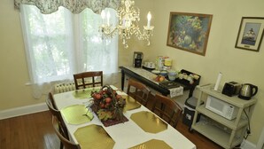 Uma sala de jantar com uma mesa posta para quatro pessoas, um candelabro, uma pintura de frutas e um carrinho de cozinha com eletrodomésticos.