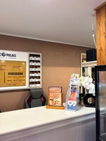 Uma receção com um painel exibindo números de quartos e tarifas, uma pequena vitrine para um menu de pequeno-almoço e um frigorífico.