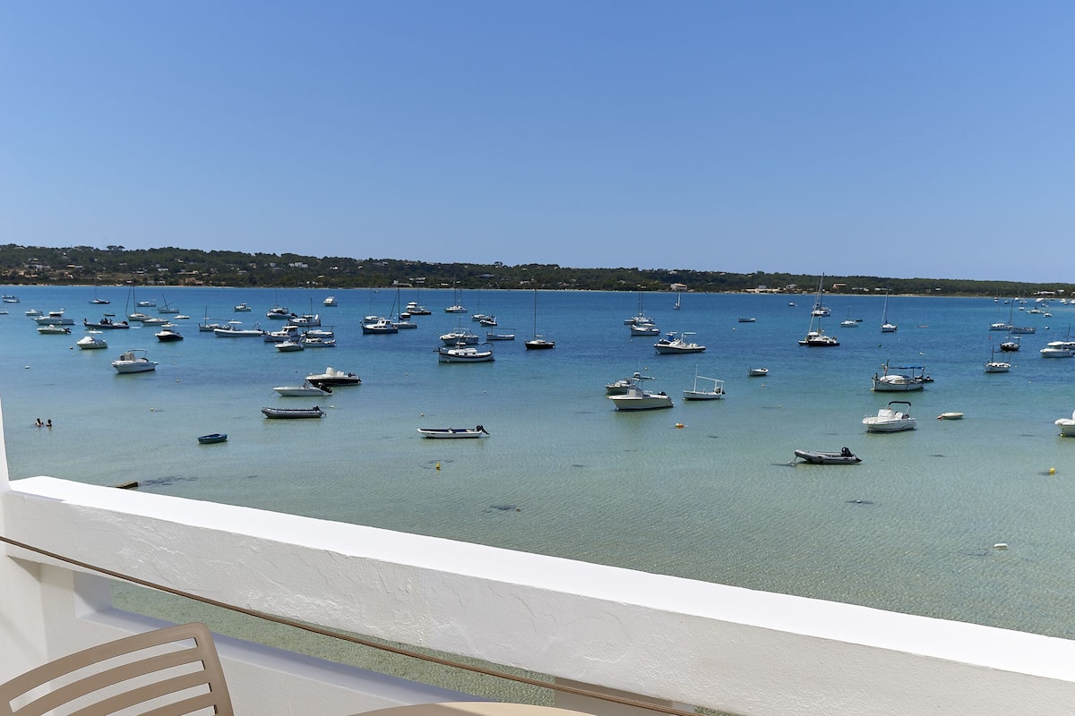 Varanda com mesa redonda e duas cadeiras, com vista para um porto cheio de barcos.