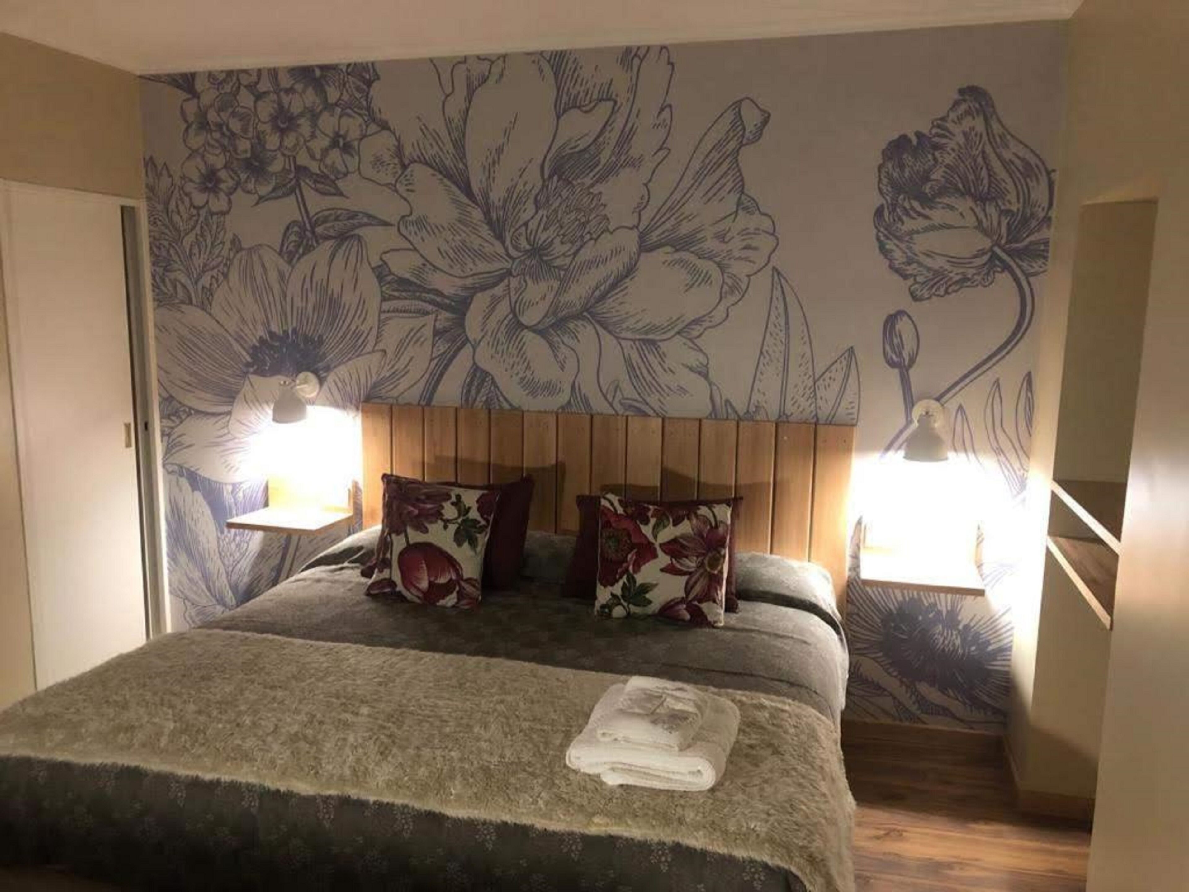 Quarto com painel floral, cabeceira de madeira, duas luminárias de mesa e uma cama bem arrumada com toalha dobrada.