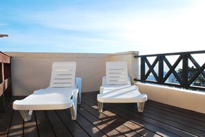 Duas poltronas brancas em um deck de madeira com guarda-corpo preto e um céu azul claro ao fundo.