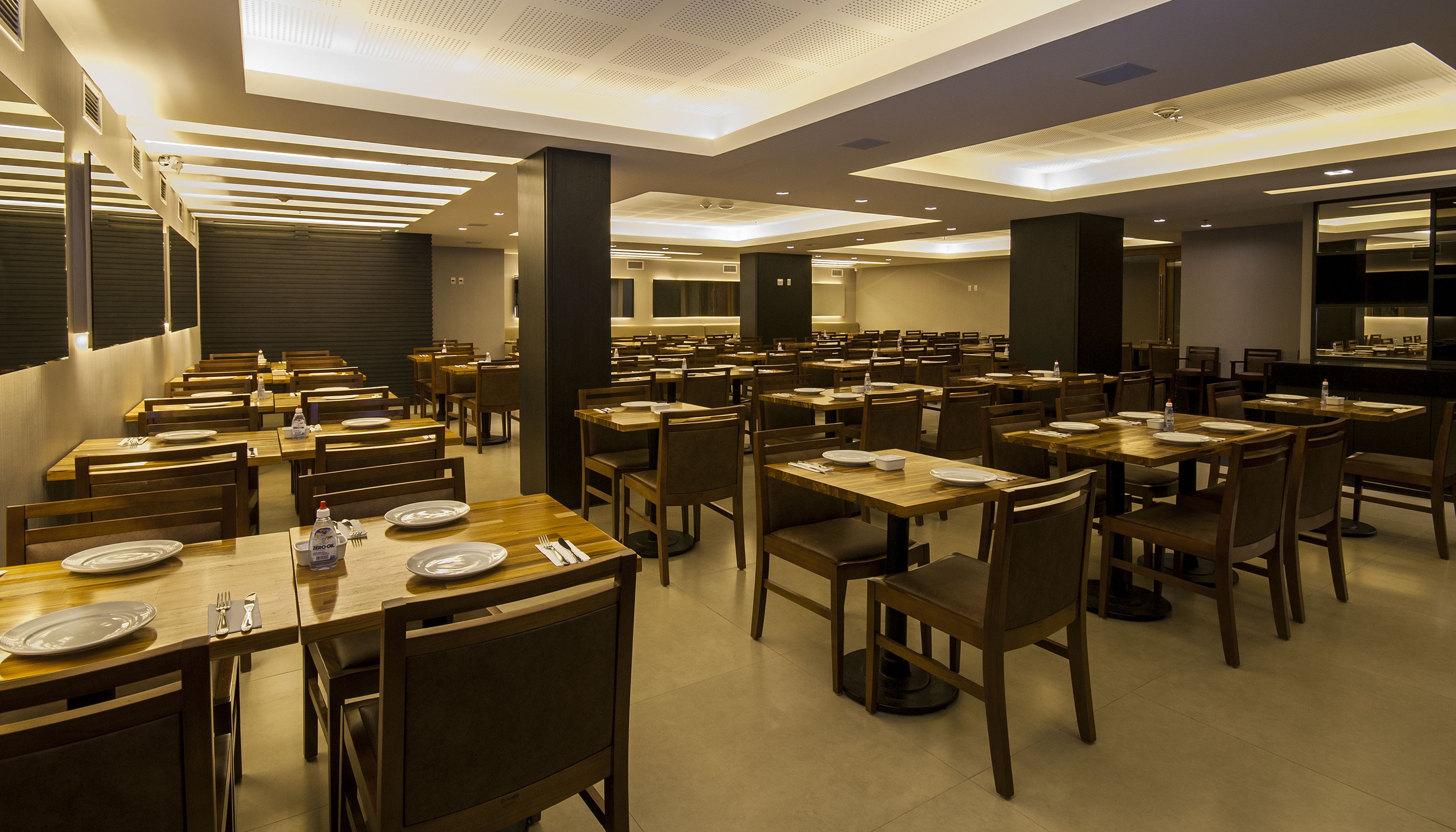 Uma ampla área de jantar com várias mesas e cadeiras, bem iluminada por luzes de teto, e um design de interiores moderno.