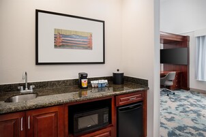 Uma cozinha de hotel com bancadas de granito, cafeteira, micro-ondas, lava-louças e um quadro na parede.