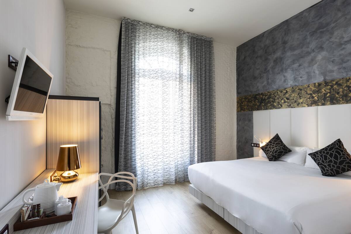 Quarto de hotel moderno com uma cama grande, televisão de tela plana na parede, mesa de cabeceira com abajur e uma cadeira.