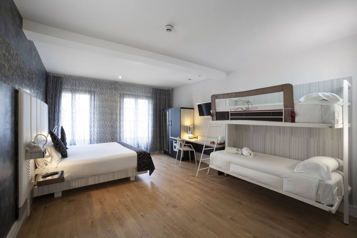 Quarto de hotel com uma cama grande, uma escrivaninha com cadeira, uma beliche e um guarda-roupa.