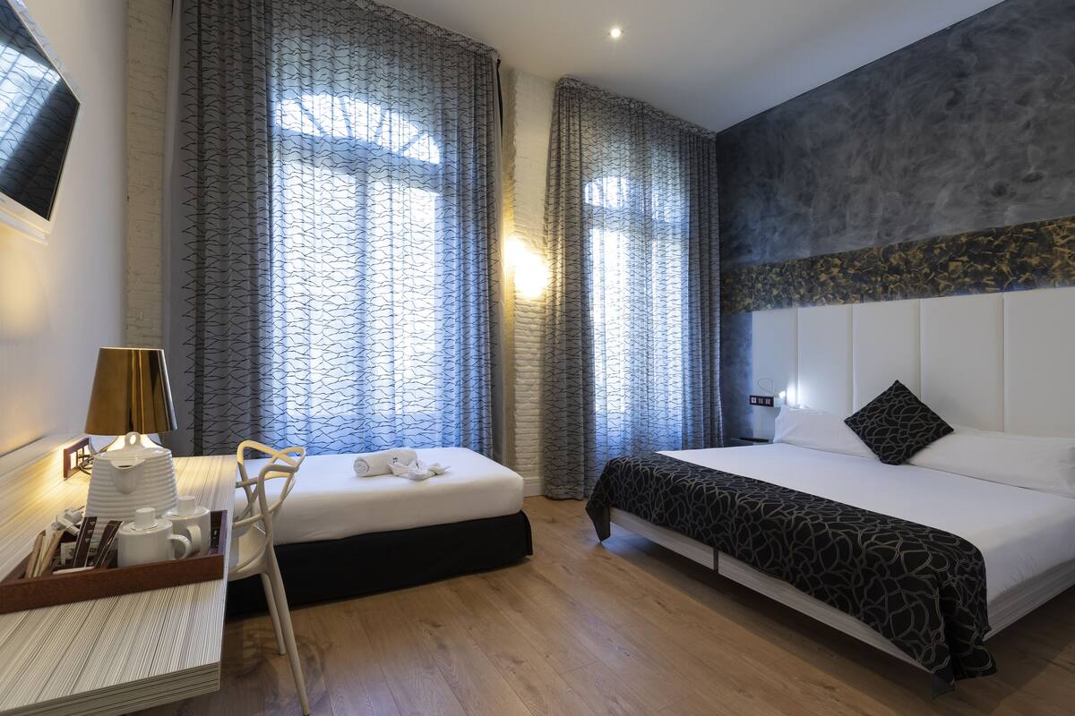 Quarto de hotel moderno com uma cama grande, uma escrivaninha com abajur e uma cadeira. Há cortinas nas janelas e uma TV fixada na parede.