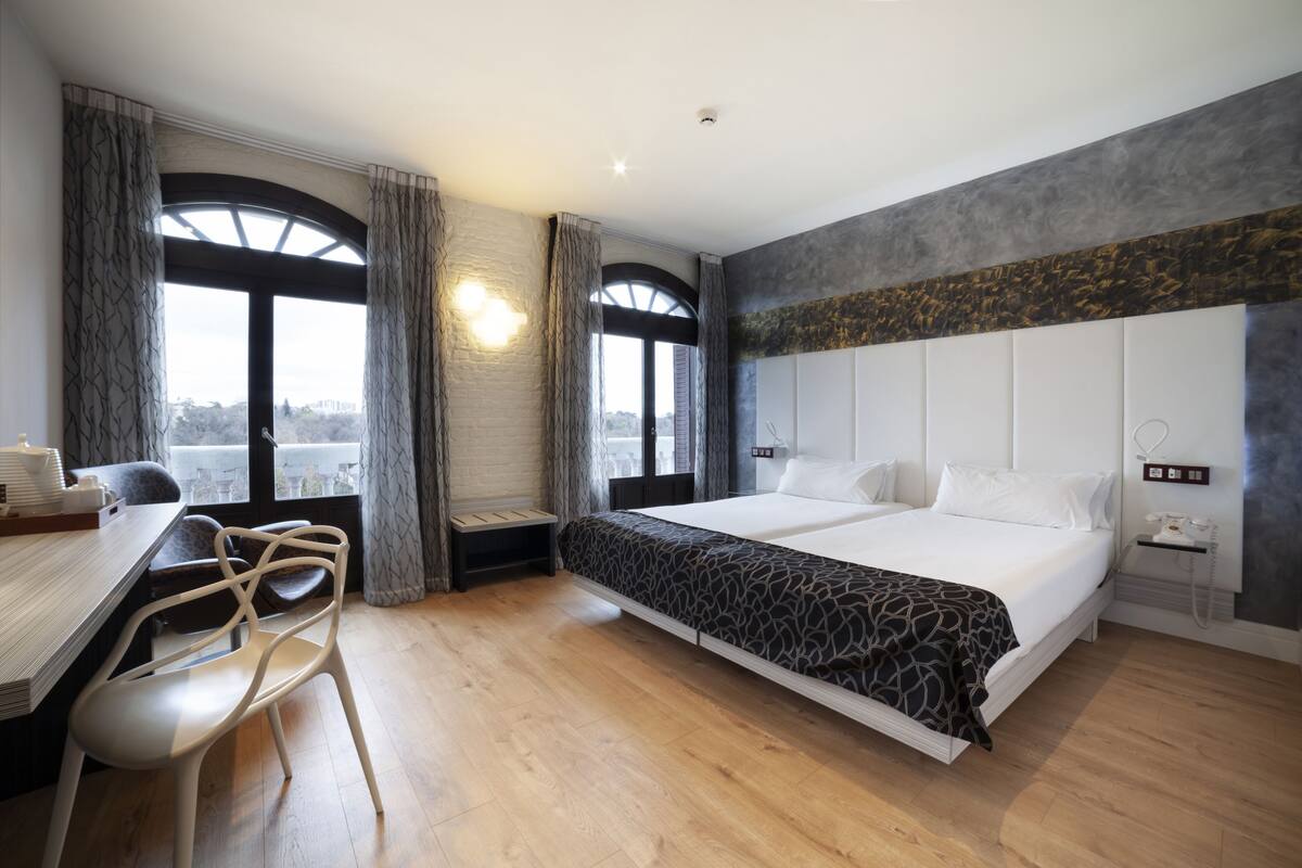 Quarto de hotel com uma cama grande, uma escrivaninha com cadeira e vista através de janelas em arco.