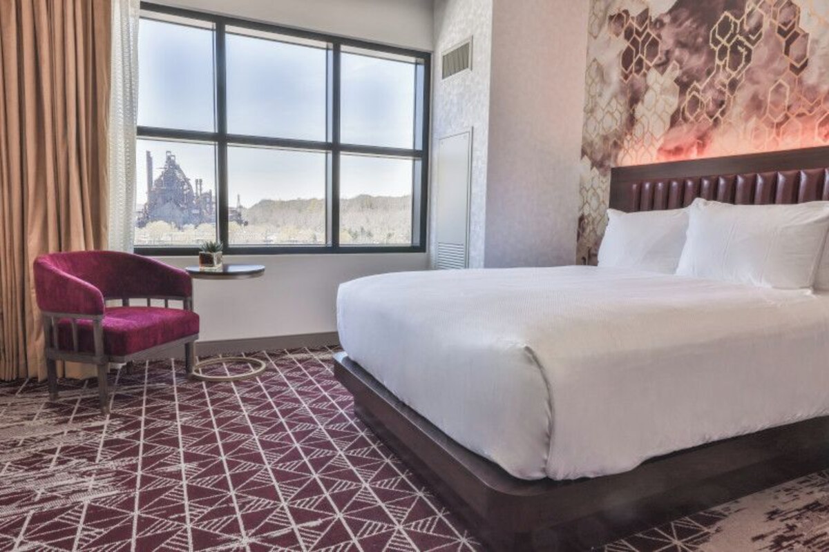 Quarto de hotel com uma cama grande, uma cadeira, uma mesinha e uma janela com vista para uma paisagem distante.