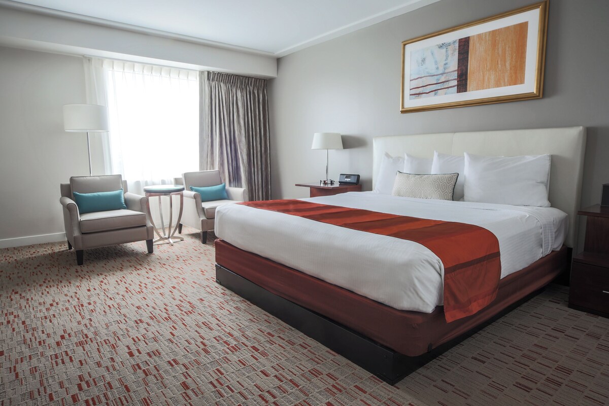 Quarto de hotel com uma cama grande, duas poltronas, uma mesa pequena, um abajur e um quadro na parede.
