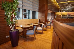 Restaurante com mobiliário de madeira, uma grande planta em vaso e teto alto com padrão em grade.