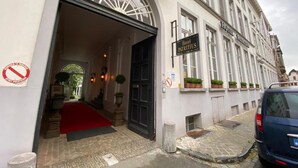 Entrada do Hotel Patrius com tapete vermelho, paredes brancas e estilo arquitetônico clássico.