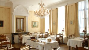 Uma sala de jantar com várias mesas postas para refeição, um candelabro e grandes janelas com cortinas.
