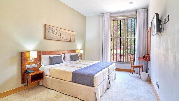 Uma cama de casal com cabeceira de madeira, mesas de cabeceira com abajures, uma janela grande com cortinas e uma TV fixada na parede.