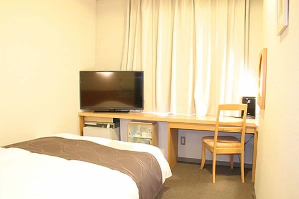 Quarto de hotel com cama, escrivaninha com televisão, cadeira e mesa pequena.