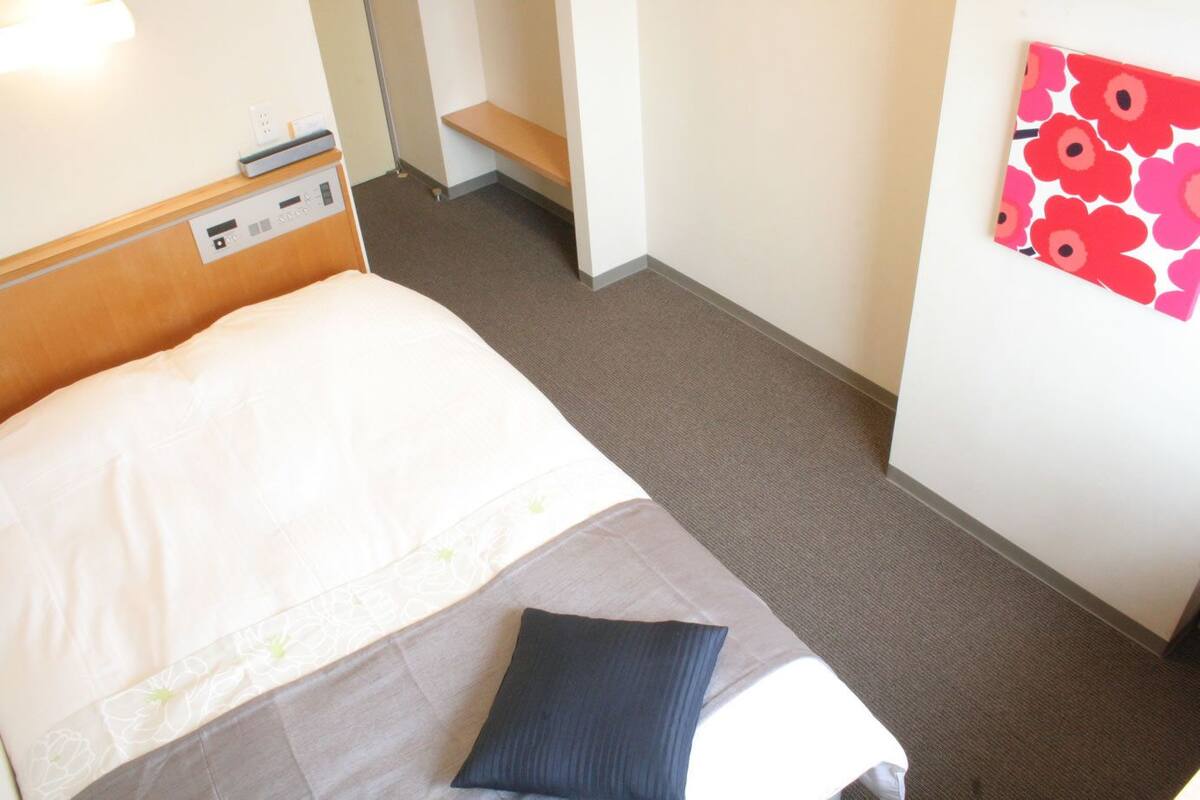 Quarto de hotel com cama, mesa de cabeceira com abajur, tomada e parede com padrão floral.