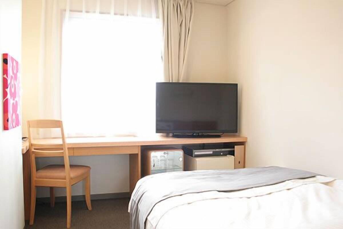 Quarto de hotel com cama, secretária com televisão, cadeira e janela com cortinas.