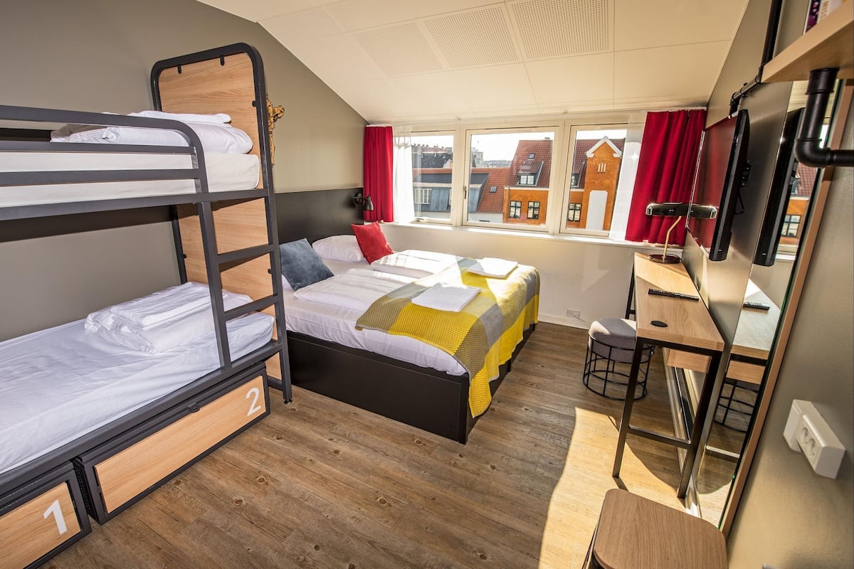 Quarto de estudante com beliches, escrivaninha e janela com vista para edifícios.