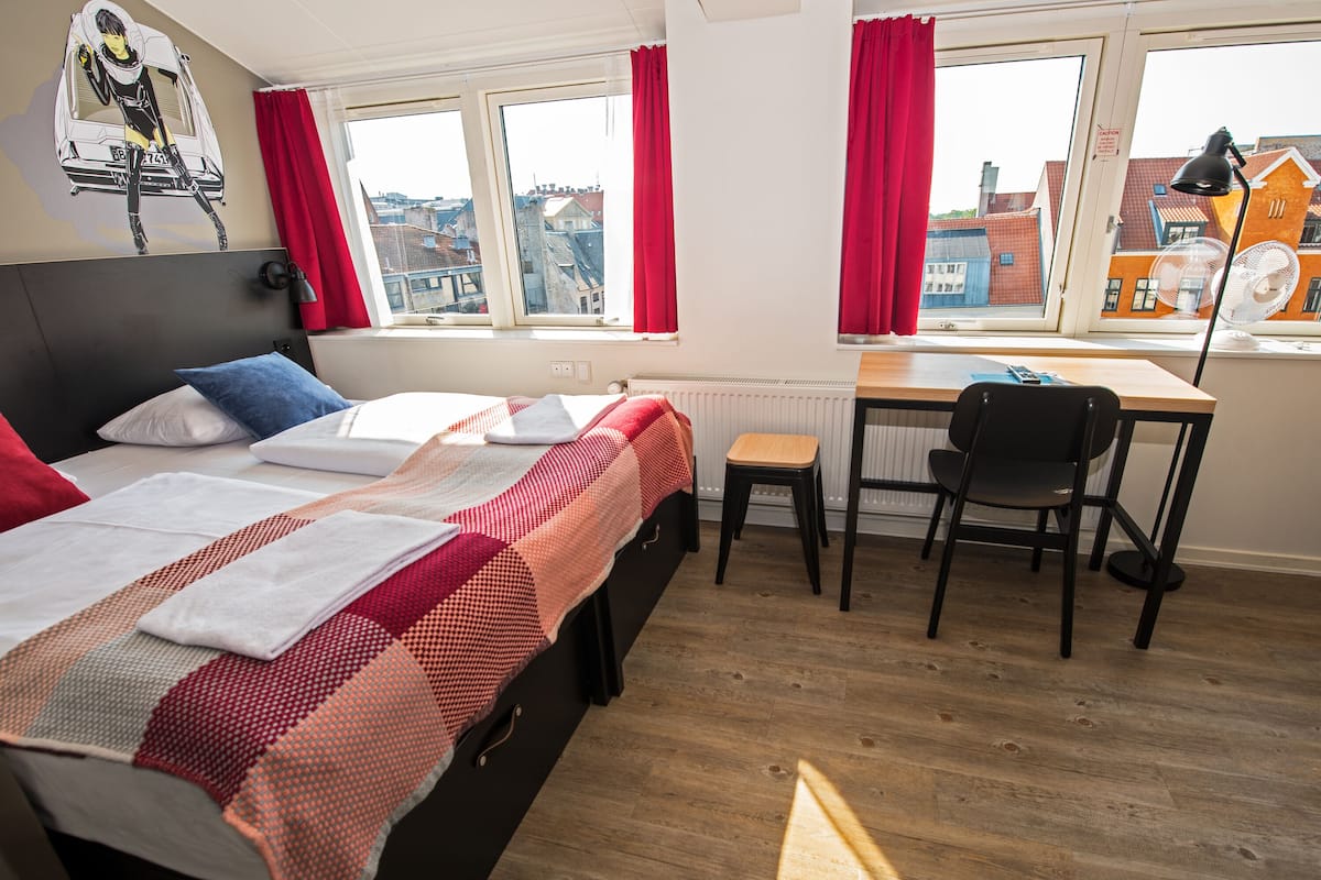 Quarto com cama, escrivaninha e cadeira. Janelas com cortinas vermelhas, radiador e luminária na parede.