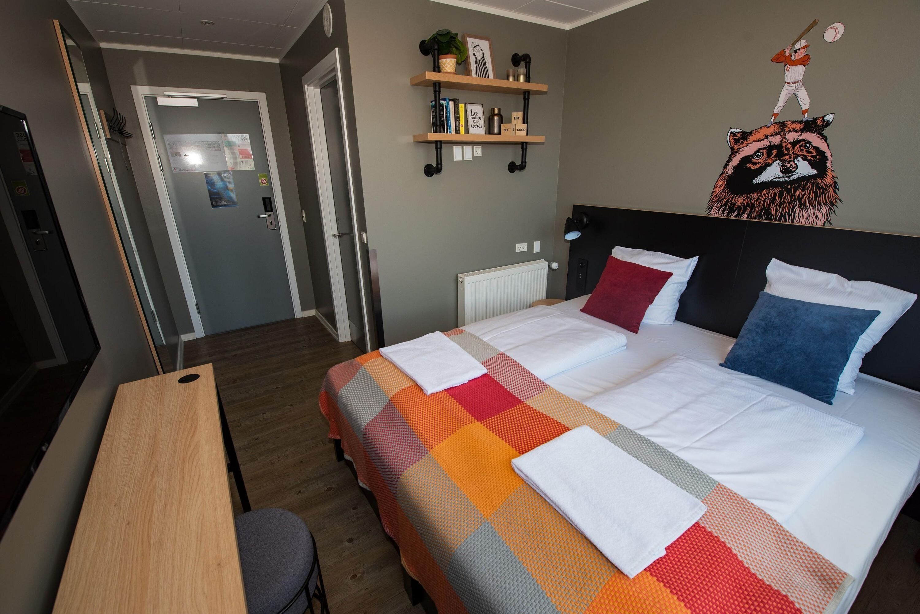 Quarto de hotel com uma cama, uma escrivaninha e uma cadeira. Há prateleiras com livros e uma planta na parede.