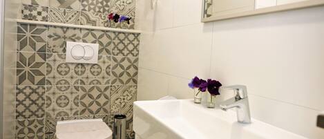 Casa de banho com azulejos estampados, sanita, bidé, lavatório com espelho e dois vasos com flores.