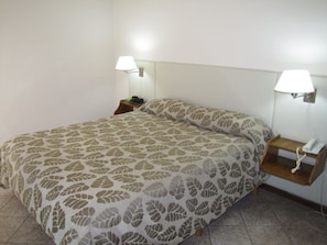 Cama bem arrumada com colcha estampada com padrão de folhas, duas luminárias de cabeceira e uma pequena mesa de cabeceira de madeira com um telefone.