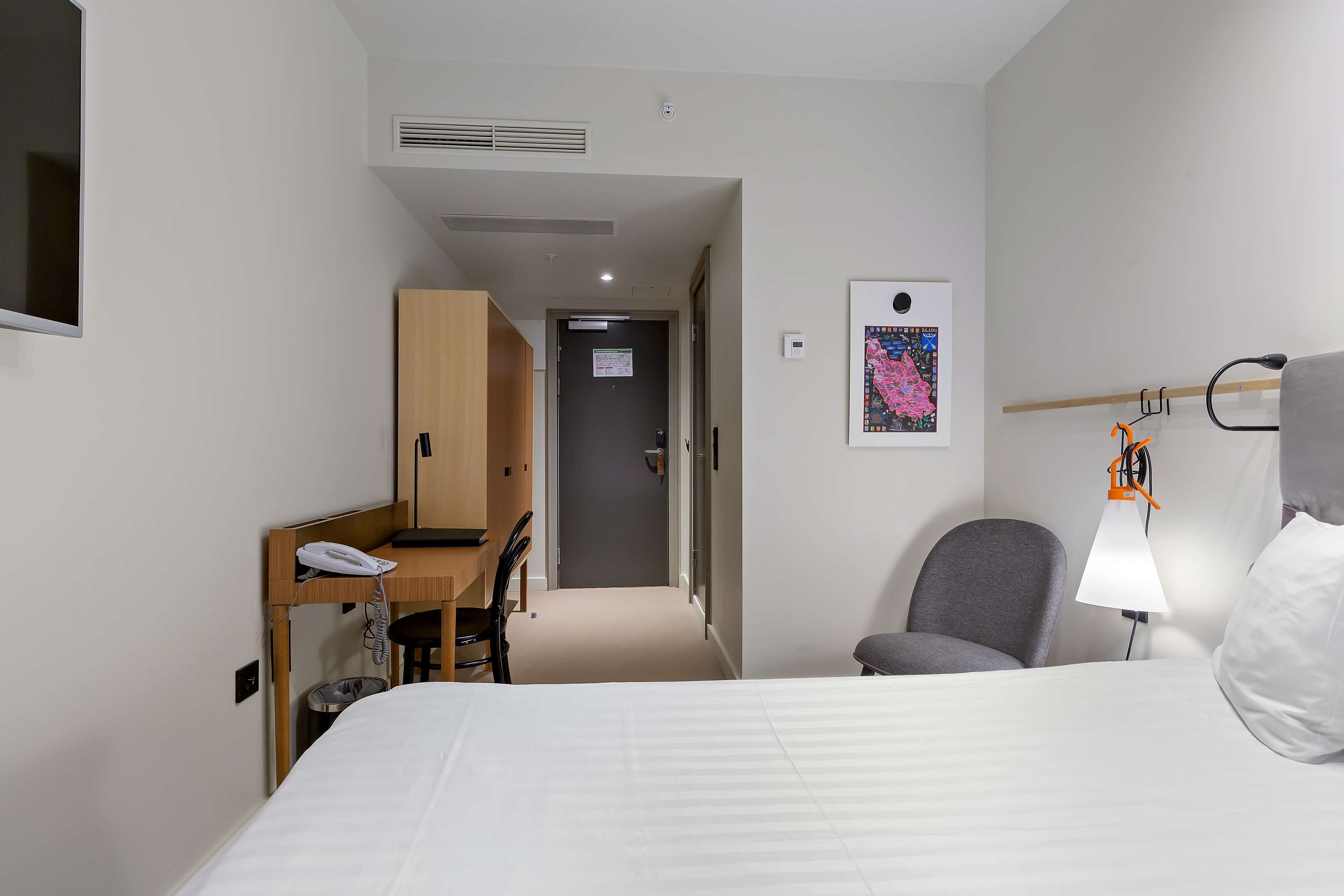 Quarto de hotel com uma cama, uma escrivaninha com telefone, uma cadeira, um abajur e uma porta que dá para outro quarto.