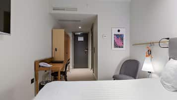 Quarto de hotel com uma cama, uma escrivaninha com telefone, uma cadeira, um abajur e uma porta que dá para outro quarto.