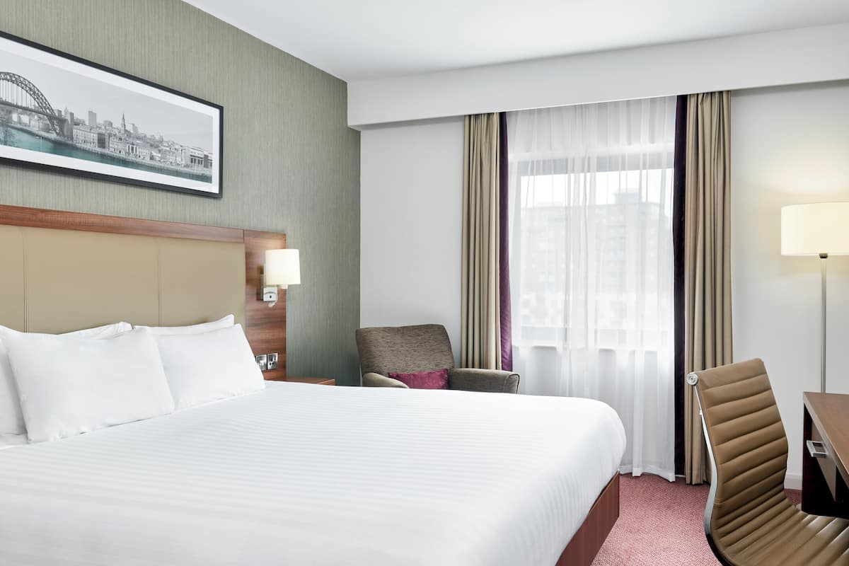 Quarto de hotel com uma cama grande, uma escrivaninha com televisão, uma cadeira, mesa de cabeceira com abajur e uma janela com cortinas.