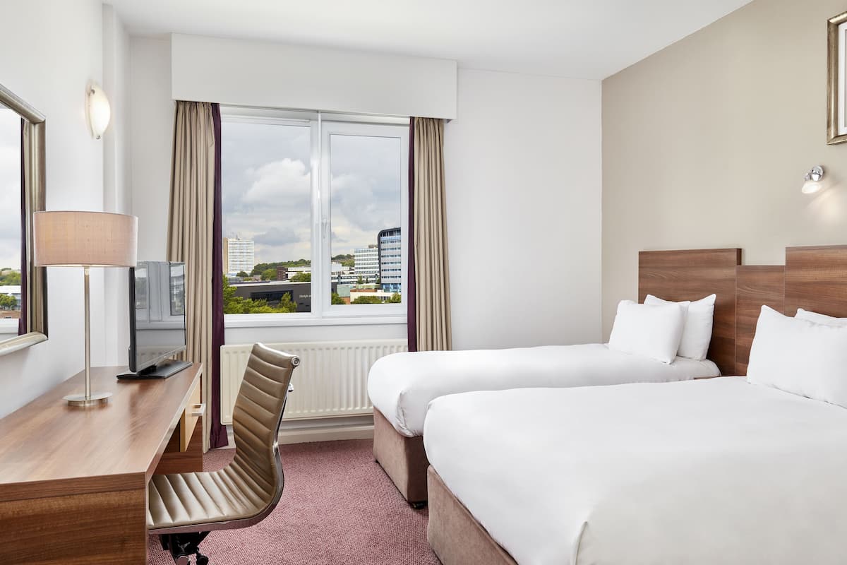 Quarto de hotel com duas camas, uma escrivaninha com abajur, uma cadeira e uma televisão.