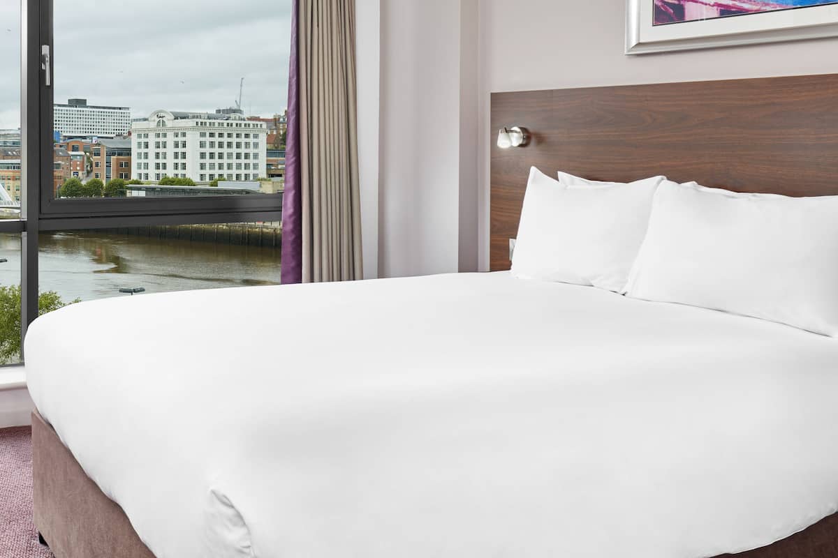 Quarto de hotel com uma cama grande, cabeceira de madeira e vista para a cidade através da janela.