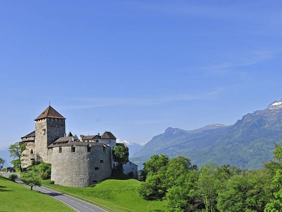 Um castelo de pedra histórico com uma torre imponente, rodeado por colinas verdejantes e montanhas ao fundo.