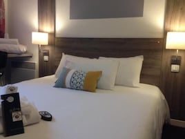 Quarto de hotel com uma cama grande, duas luminárias de cabeceira, um criado-mudo e uma televisão fixada na parede.