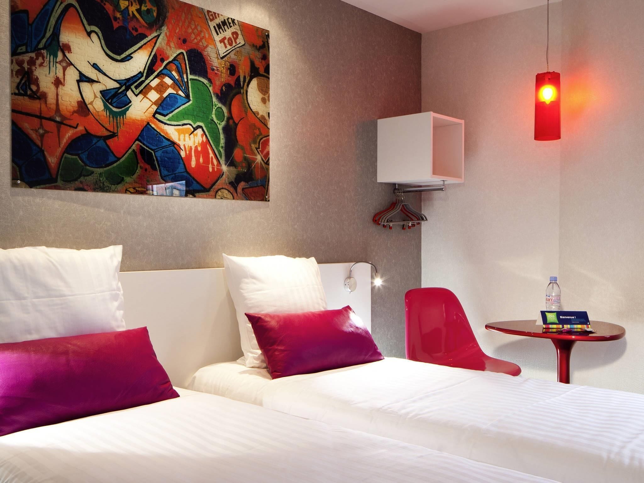 Quarto de hotel com cama, almofadas, uma cadeira vermelha, uma mesinha com uma garrafa e parede com arte de graffiti.