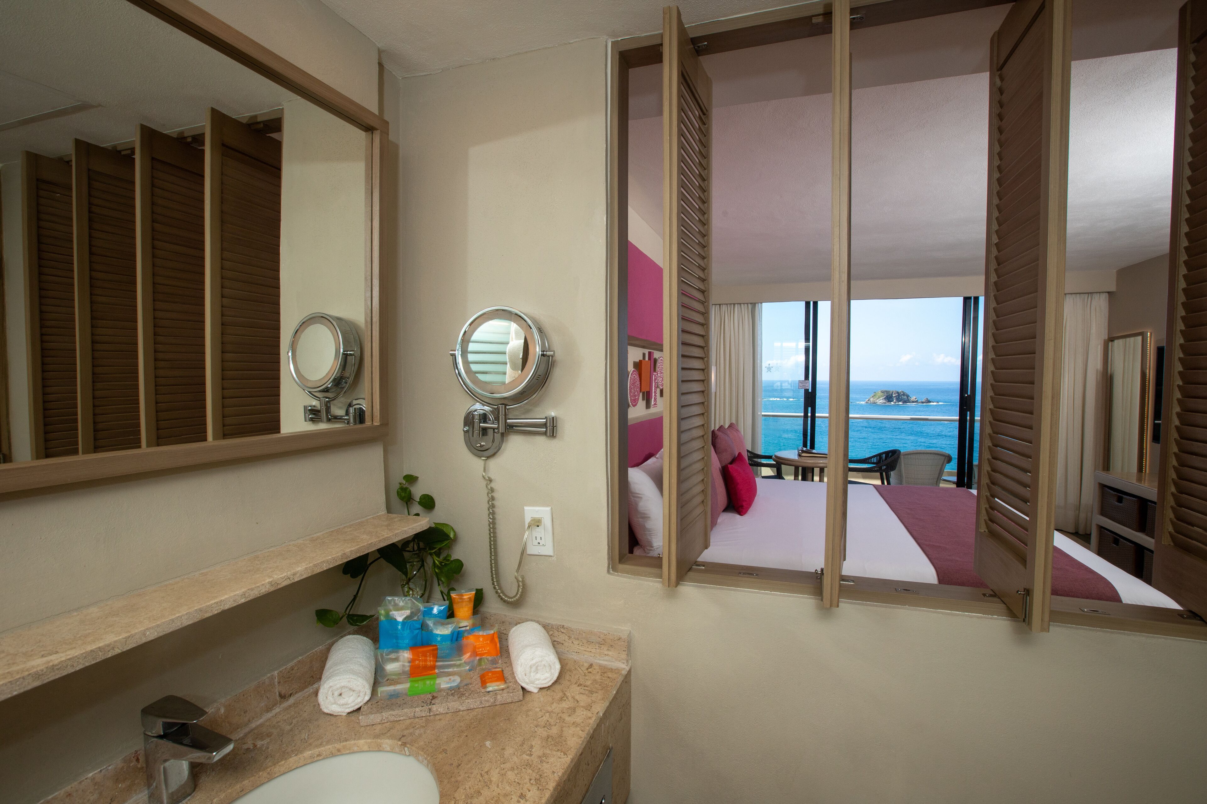 Casa de banho de hotel com lavatório, espelhos e artigos de higiene. Tem vista para o mar através de uma porta aberta.