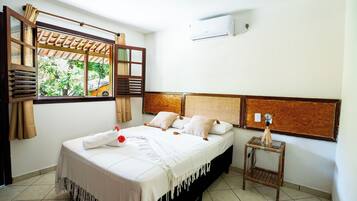 Quarto de hotel com cama, janela com cortinas, ar condicionado fixado na parede e uma pequena mesa com luminária.