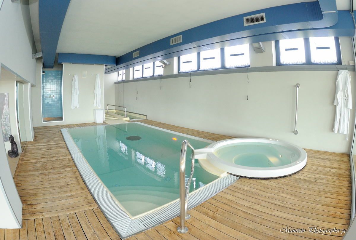 Uma grande piscina interior com um deck de madeira, uma banheira de hidromassagem e vista para a cidade através de grandes janelas.
