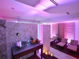 Uma área de spa moderna com parede de pedra, bacia de metal, garrafas e uma cesta de frutas. Há duas cadeiras e uma mesa com velas.
