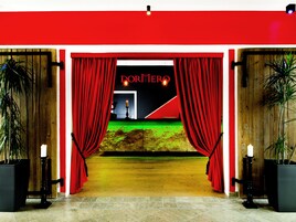 Entrada de teatro com cortinas vermelhas, palco com piso verde e um letreiro com a palavra 'DORNER'.