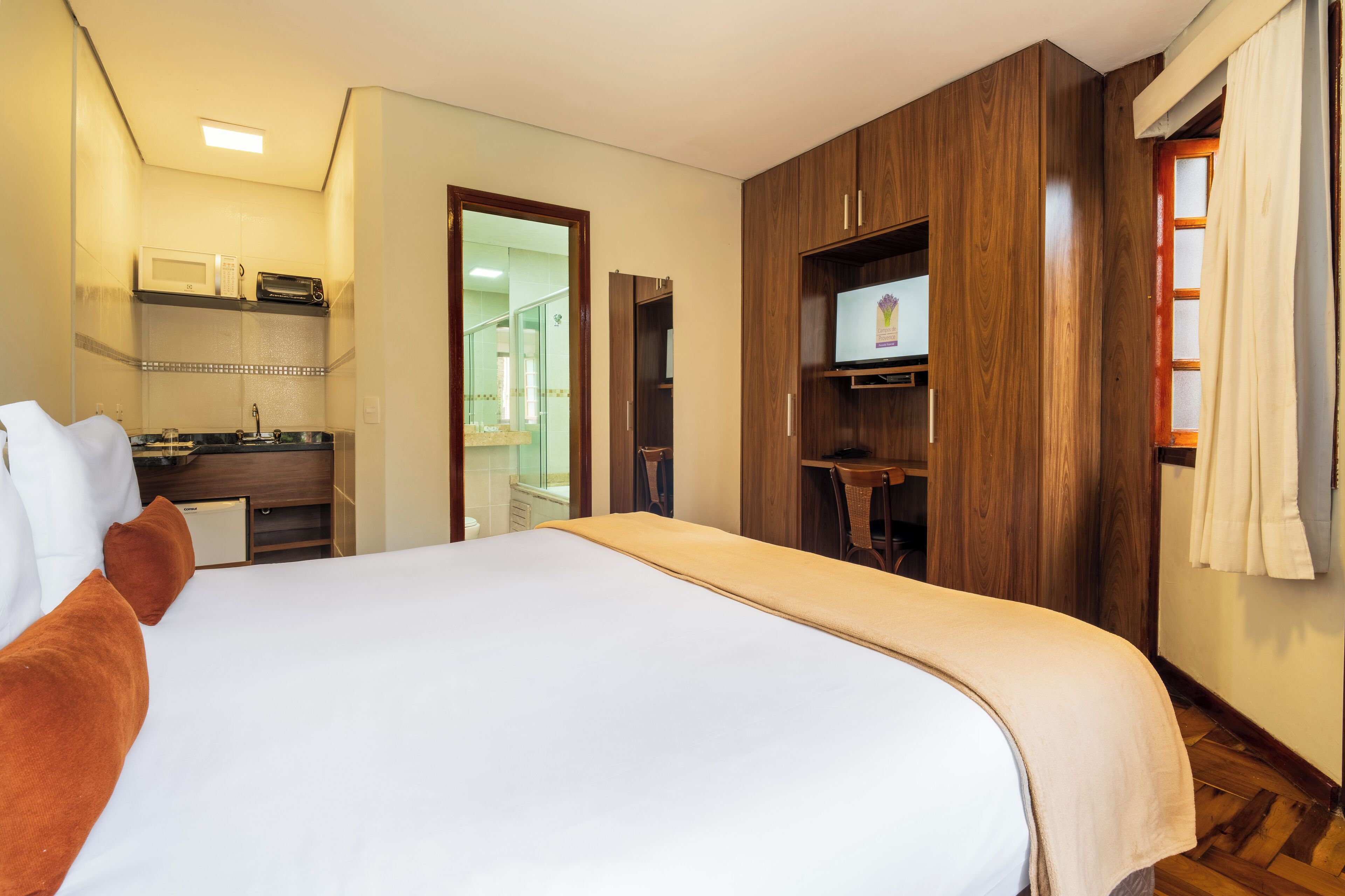 Quarto de hotel com uma cama grande, guarda-roupa de madeira, pequeno espaço de cozinha e uma televisão.