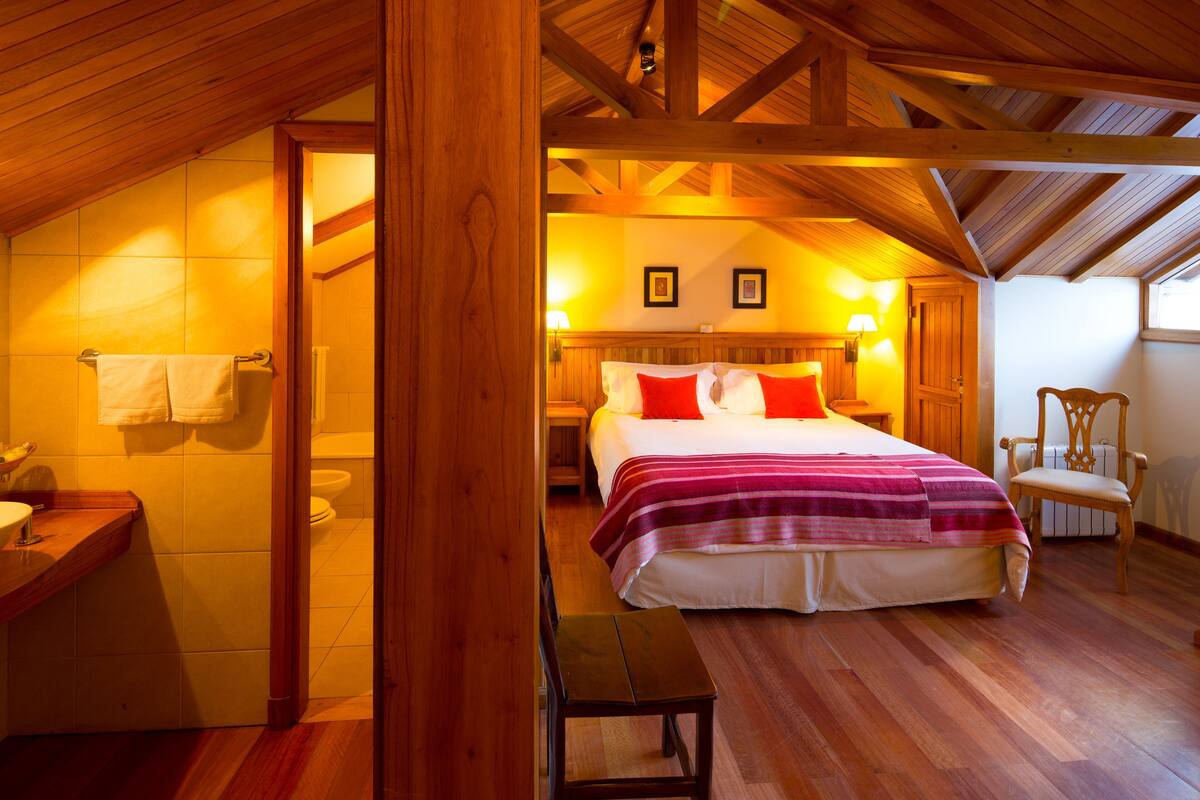 Um quarto aconchegante com teto de madeira, cama com manta listrada, cadeira de madeira e mesa de cabeceira com abajur.