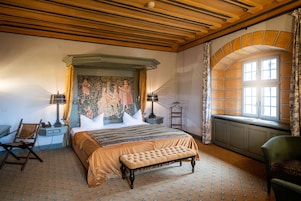 Quarto com uma cama grande, um banquinho, uma cadeira, uma mesa de cabeceira, um abajur, uma tapeçaria e uma janela com cortinas.