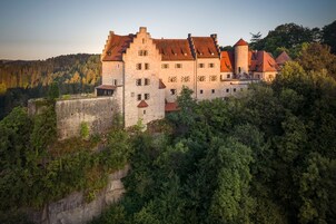 Um castelo histórico com várias torres e uma muralha fortificada, rodeado por uma floresta densa.