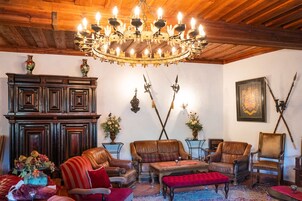 Quarto tradicional com teto de madeira, um grande candelabro, poltronas de couro, um armário de madeira e um brasão emoldurado na parede.