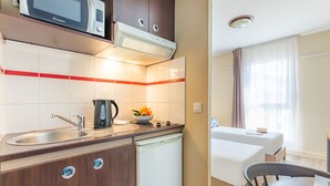 Cozinha compacta com micro-ondas, pia e pequena geladeira. No quarto adjacente, há uma cama com mesa de cabeceira e uma cadeira.