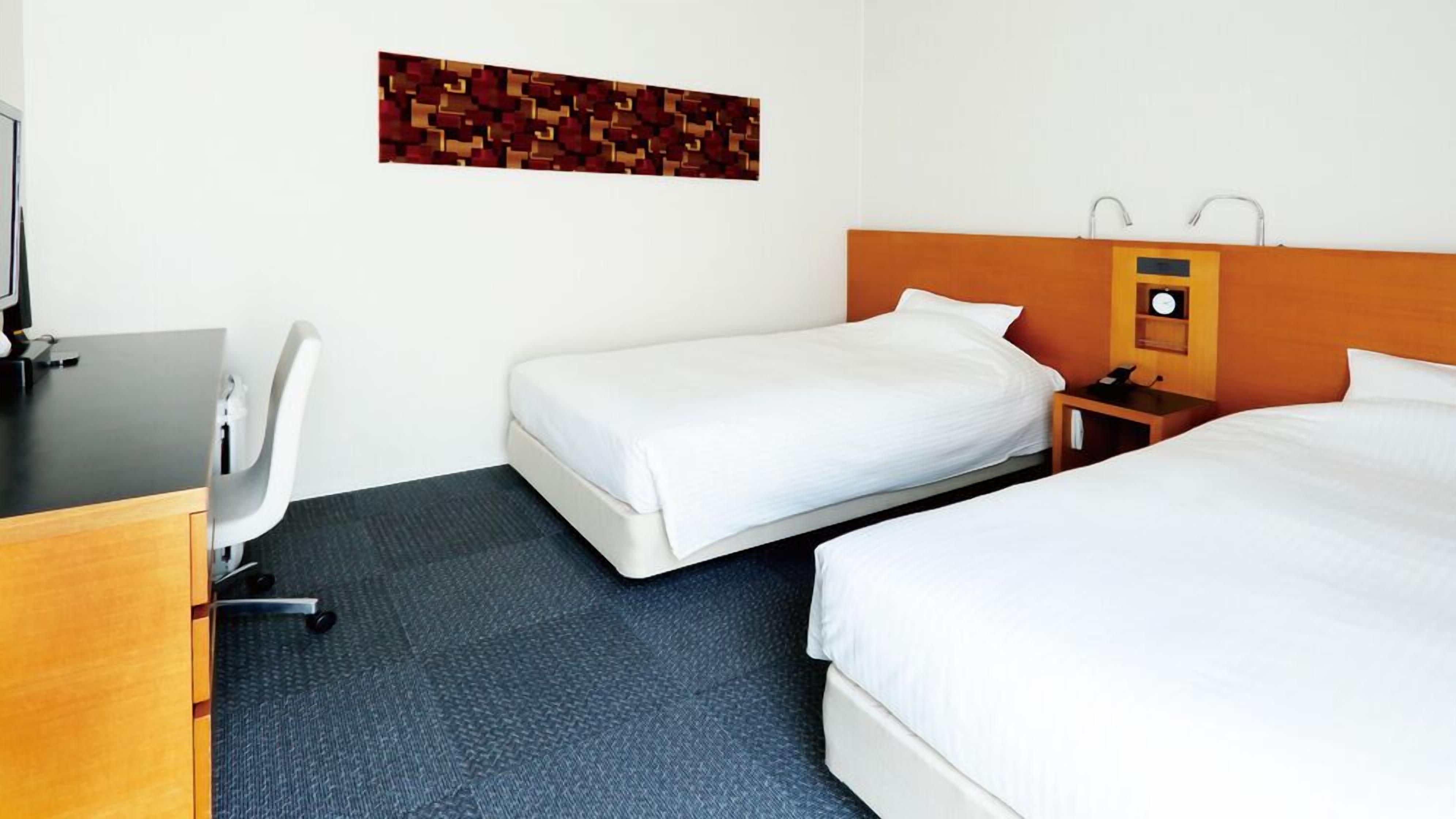 Quarto de hotel com duas camas, uma escrivaninha, uma cadeira, uma mesinha de centro com um relógio e uma obra de arte na parede.
