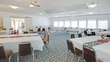 Uma ampla sala de banquetes com várias mesas e cadeiras, teto branco com vigas aparentes e carpete estampado.