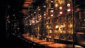 Um bar com várias prateleiras de garrafas e copos, um balcão comprido e algumas cadeiras.