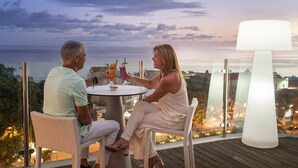 Duas pessoas sentadas à mesa num terraço, com vista para o mar e luzes da cidade.