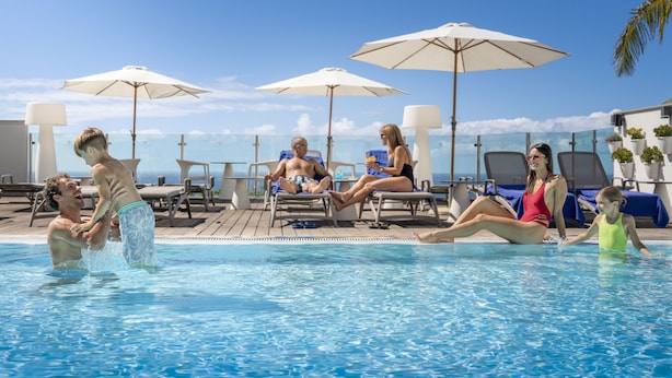 Área de piscina com espreguiçadeiras e guarda-sóis, céu claro e vista para o oceano.