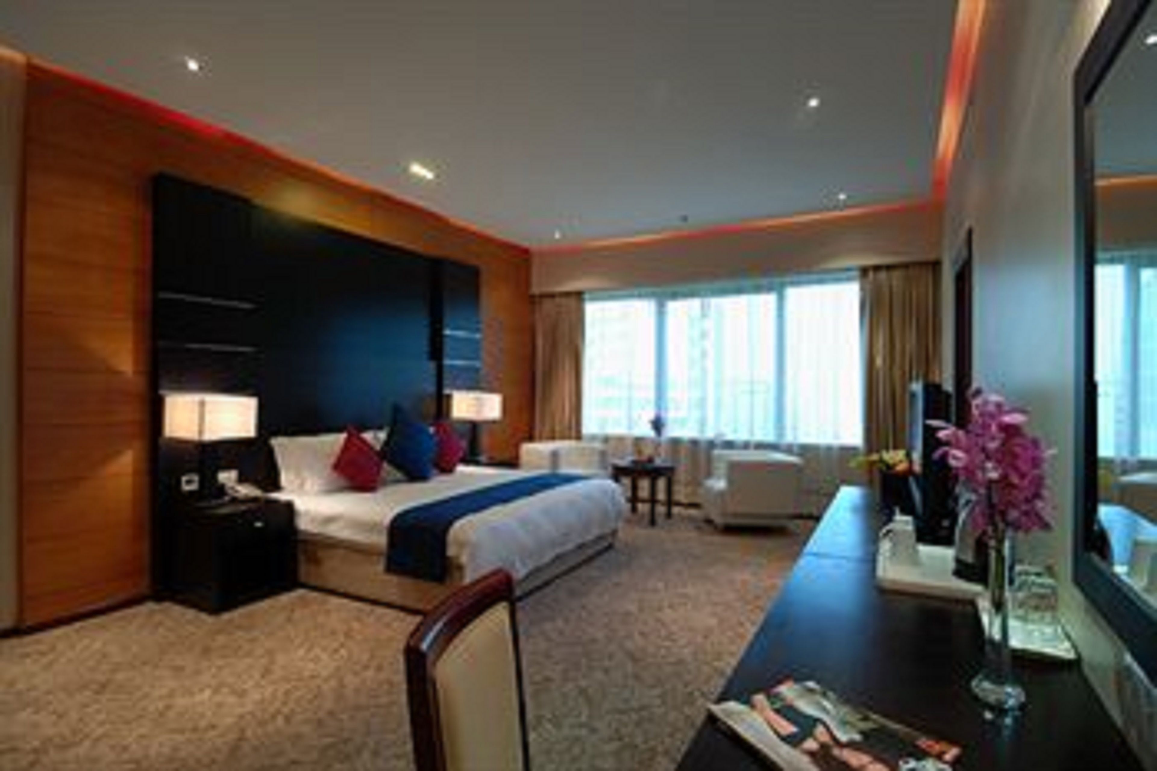 Quarto de hotel moderno com uma cama grande, uma escrivaninha com luminária, uma cadeira e uma janela ampla.